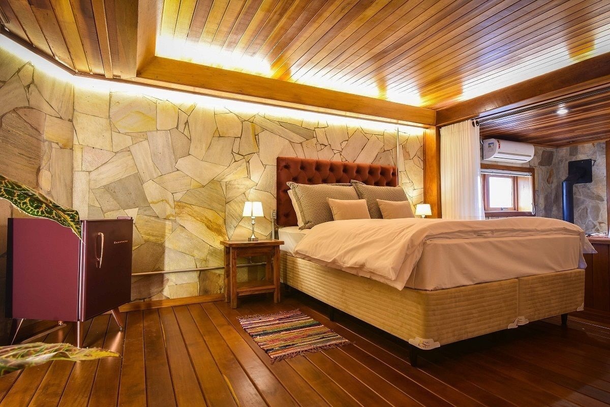 Quarto com parede de pedra, teto de madeira, cama com cabeceira, mesas de cabeceira e radiador.