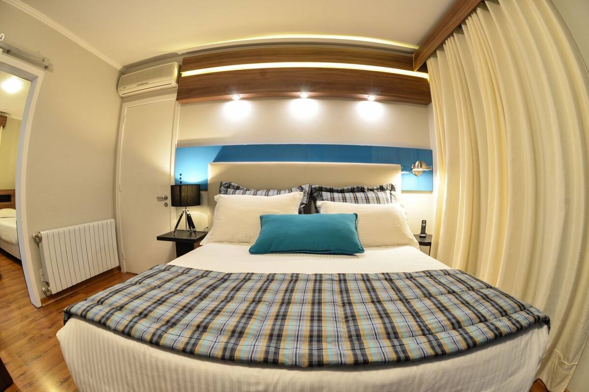 Quarto de hotel com uma cama grande, uma mesa de cabeceira com luminária, ar condicionado fixado na parede e uma janela com cortinas.