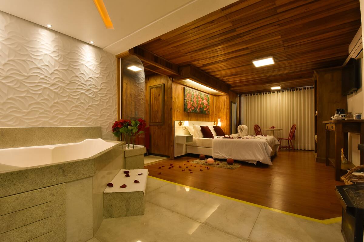 Um banheiro moderno com banheira, chuveiro e pia. Há uma cama com colcha e travesseiros brancos, uma mesinha com um vaso de flores e uma cadeira. O quarto tem piso e teto de madeira.