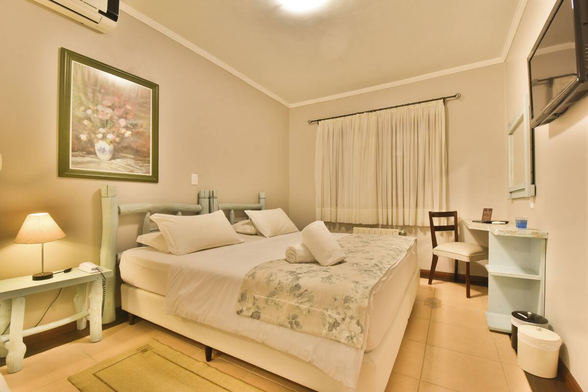 Quarto com cama, poltrona, escrivaninha, abajur, quadro e janela com cortinas.