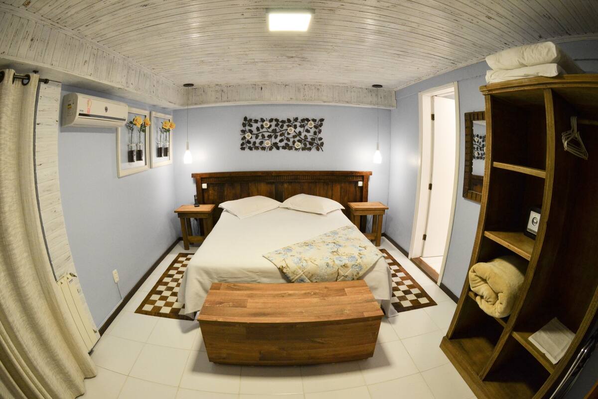 Quarto com cama de madeira, um banquinho, guarda-roupa e piso xadrez.