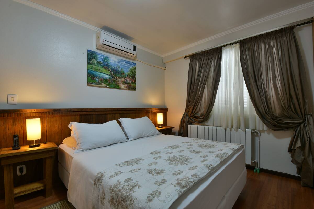 Quarto de hotel com cama, mesa de cabeceira, aparelho de ar condicionado, quadro e cortinas.
