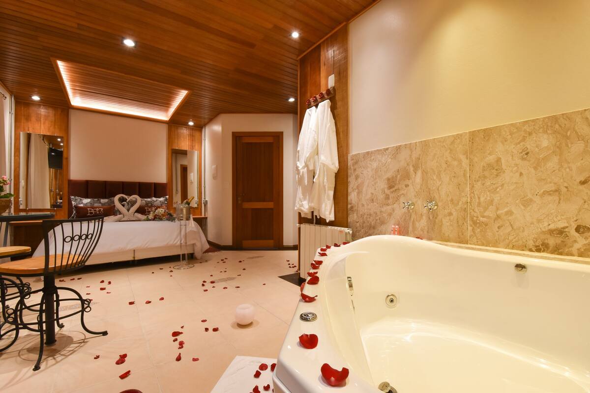 Quarto de hotel com teto de madeira, uma banheira grande, uma cama com travesseiro em formato de coração e uma cadeira.