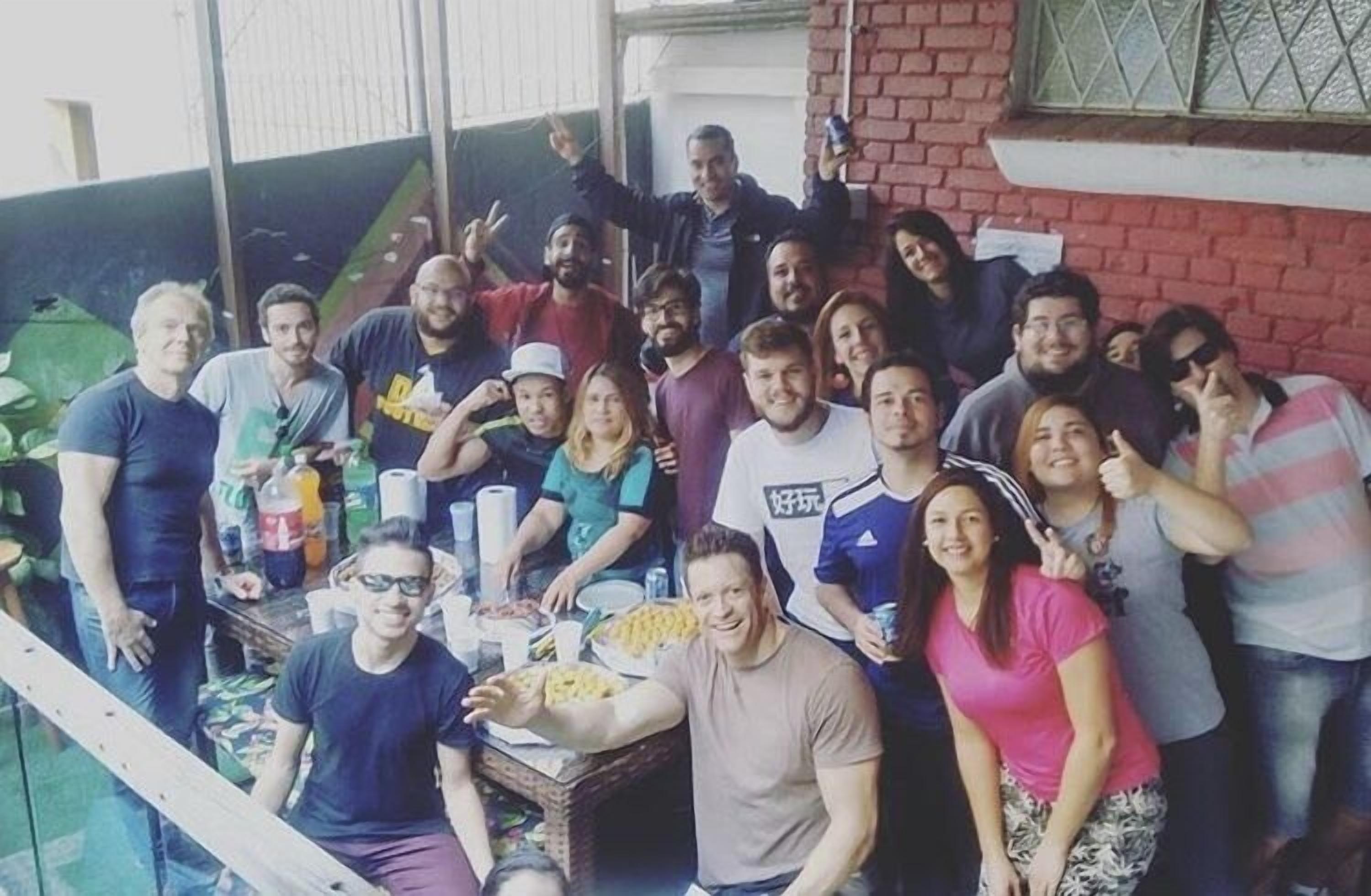 Um grupo de pessoas reunido em torno de uma mesa com comida e bebidas, em ambiente externo com parede de tijolos e janela ao fundo.