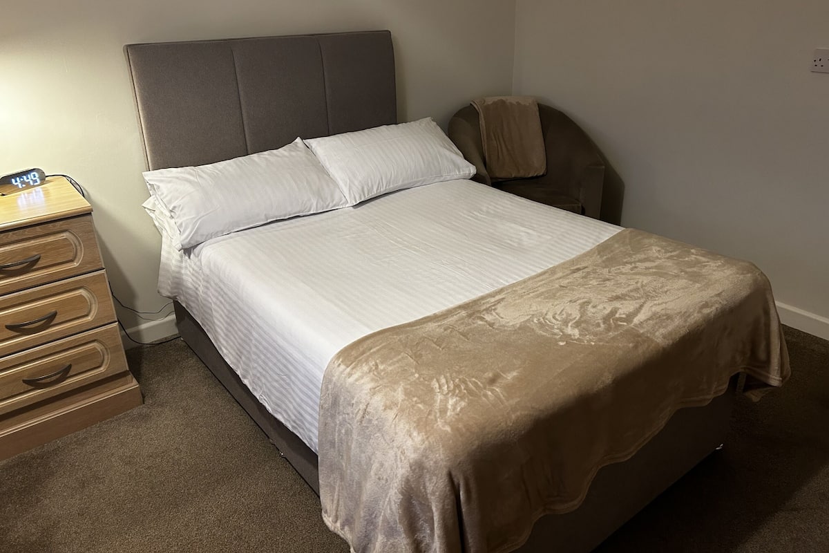 Uma cama bem arrumada com cabeceira cinzenta, uma mesa de cabeceira com gavetas e um relógio marcando 4:49.
