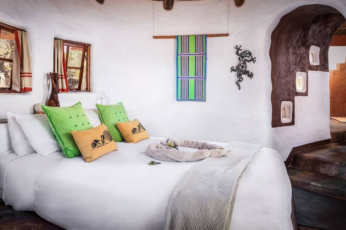 Quarto com cama, travesseiros, mesa de cabeceira, janela e uma obra de arte na parede.