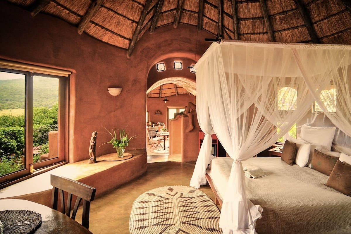 Quarto com cama de dossel, vista para o exterior, área de jantar e uma pequena estátua.