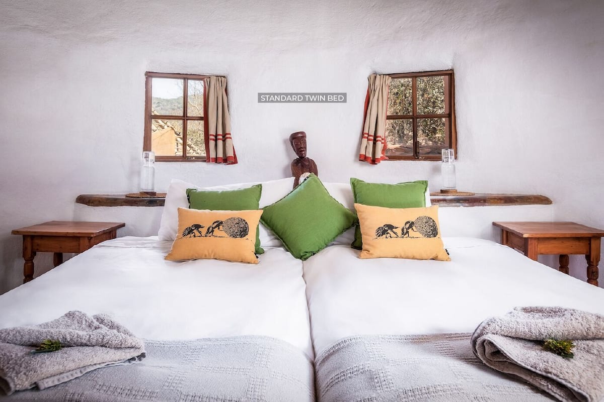 Uma cama de casal standard com duas almofadas, cabeceira de madeira e um letreiro sobre a cama. Há duas janelas com cortinas, um criado-mudo de madeira e uma mesinha.