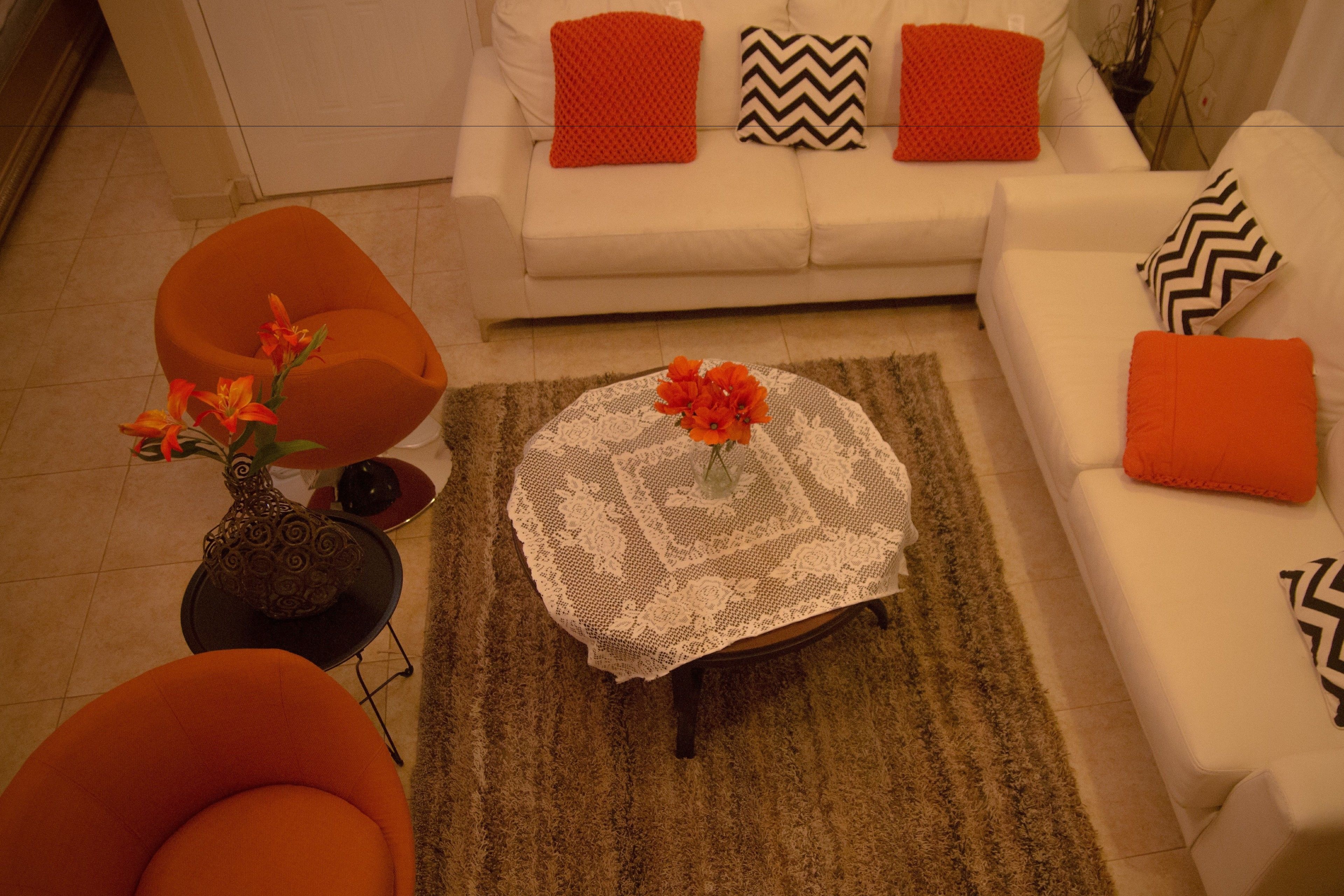 Uma sala de estar com um sofá branco, poltronas laranja, uma mesa de centro com toalha de renda e um vaso com flores.