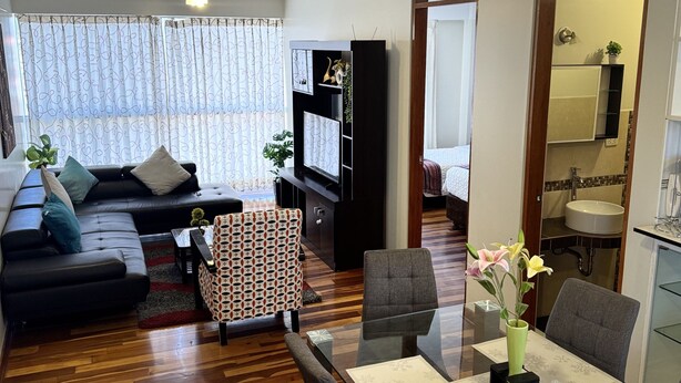 Uma sala de estar moderna com sofá, um banquinho estampado, mesa de jantar com cadeiras e área de cozinha visível por uma porta aberta.