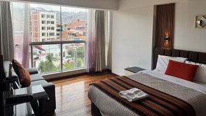 Quarto de hotel com uma janela grande, uma cama com almofadas brancas e vermelhas, uma escrivaninha com teclado e chão de madeira.