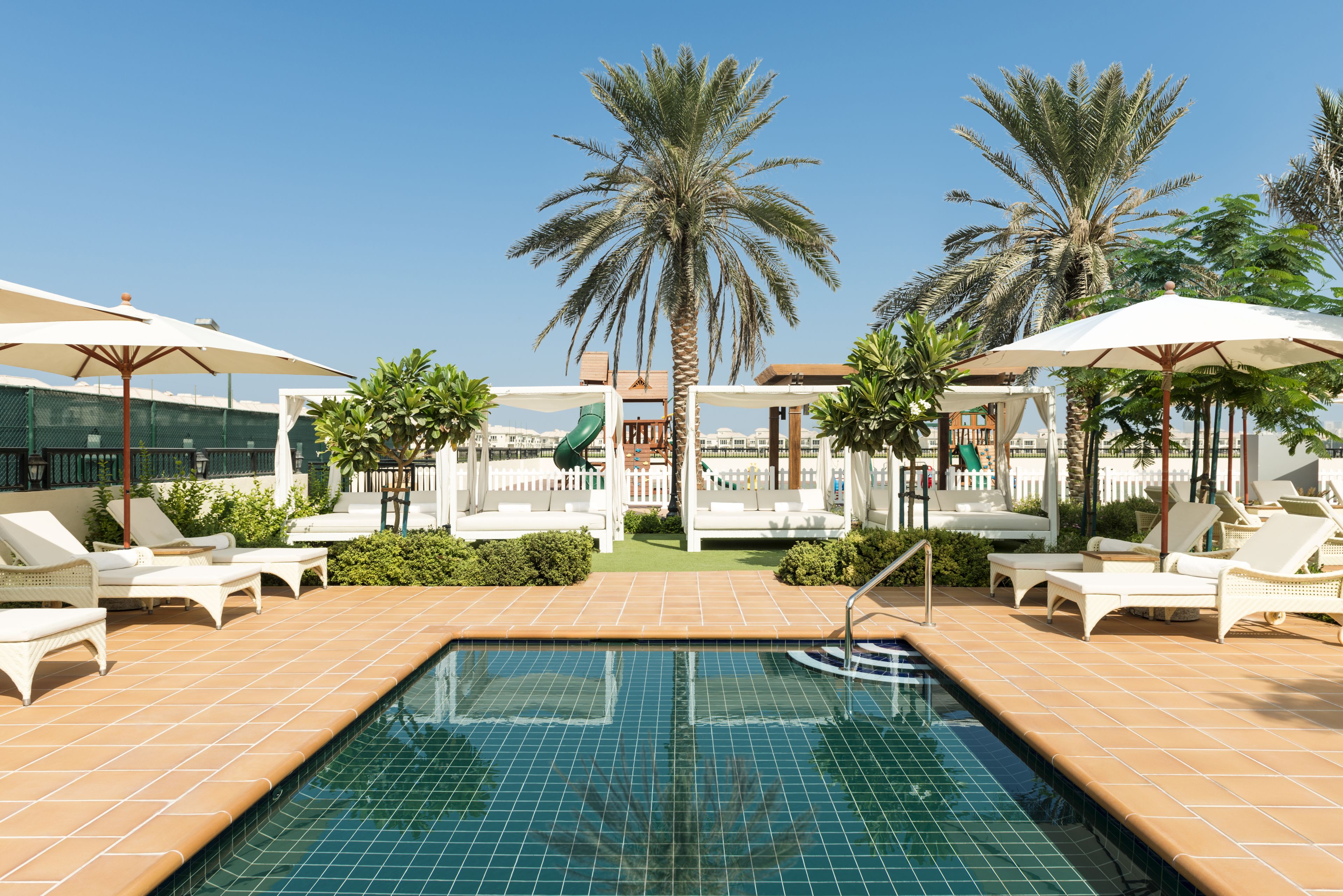 Área de piscina com espreguiçadeiras e guarda-sóis, cercada por palmeiras e um muro branco.