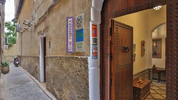 Uma rua estreita com a entrada de um edifício, apresentando uma porta de madeira com grade de metal. Há placas na parede, incluindo uma com a inscrição 'La Clef Verte 2019'.