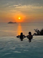 Um pôr do sol sobre um mar calmo, com duas pessoas em uma piscina infinita.