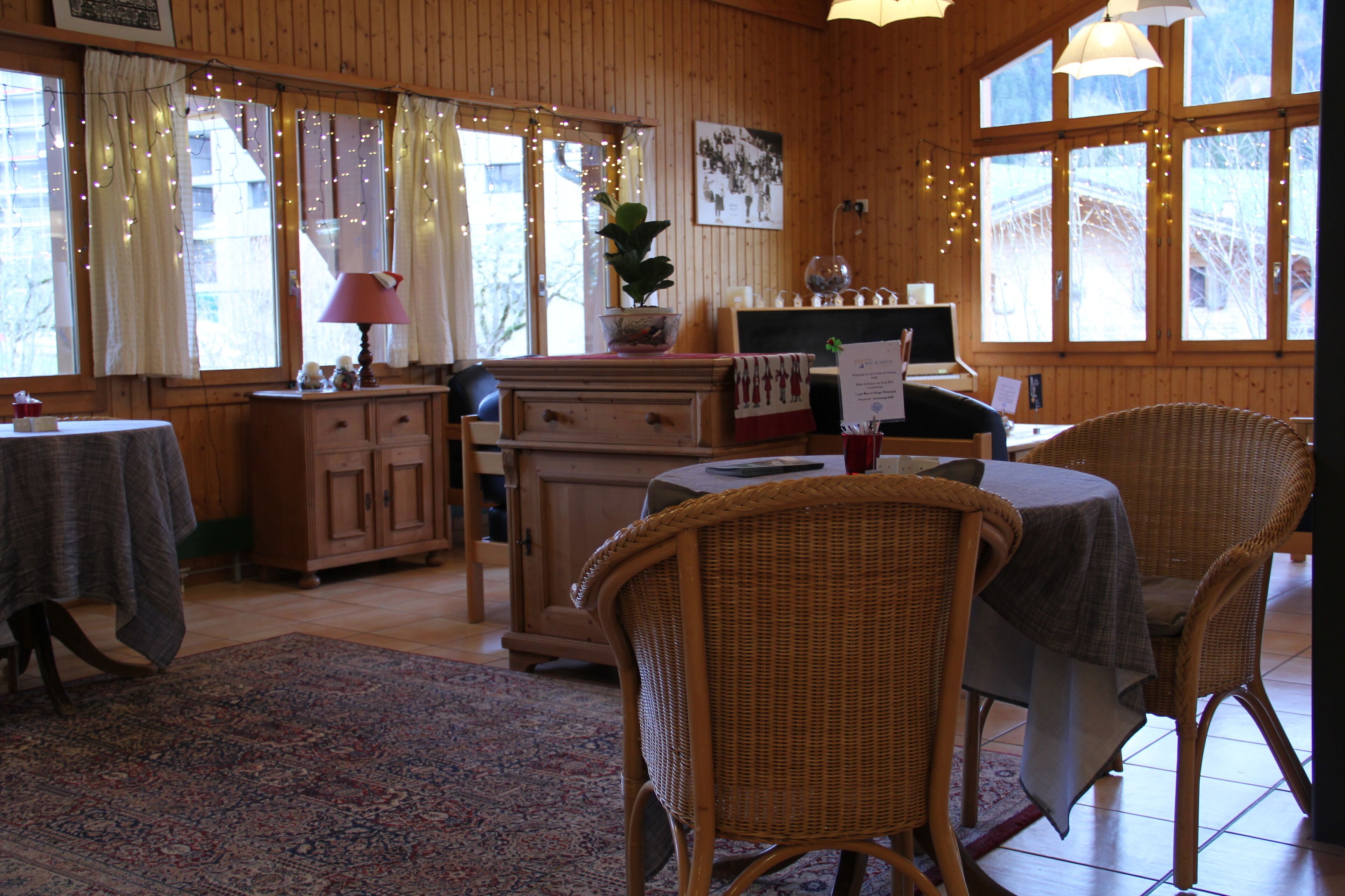 Uma área de refeições aconchegante com cadeiras de vime, uma mesa e um tapete. Há uma janela com cortinas e luzes de fada, e uma pequena planta sobre uma mesa.