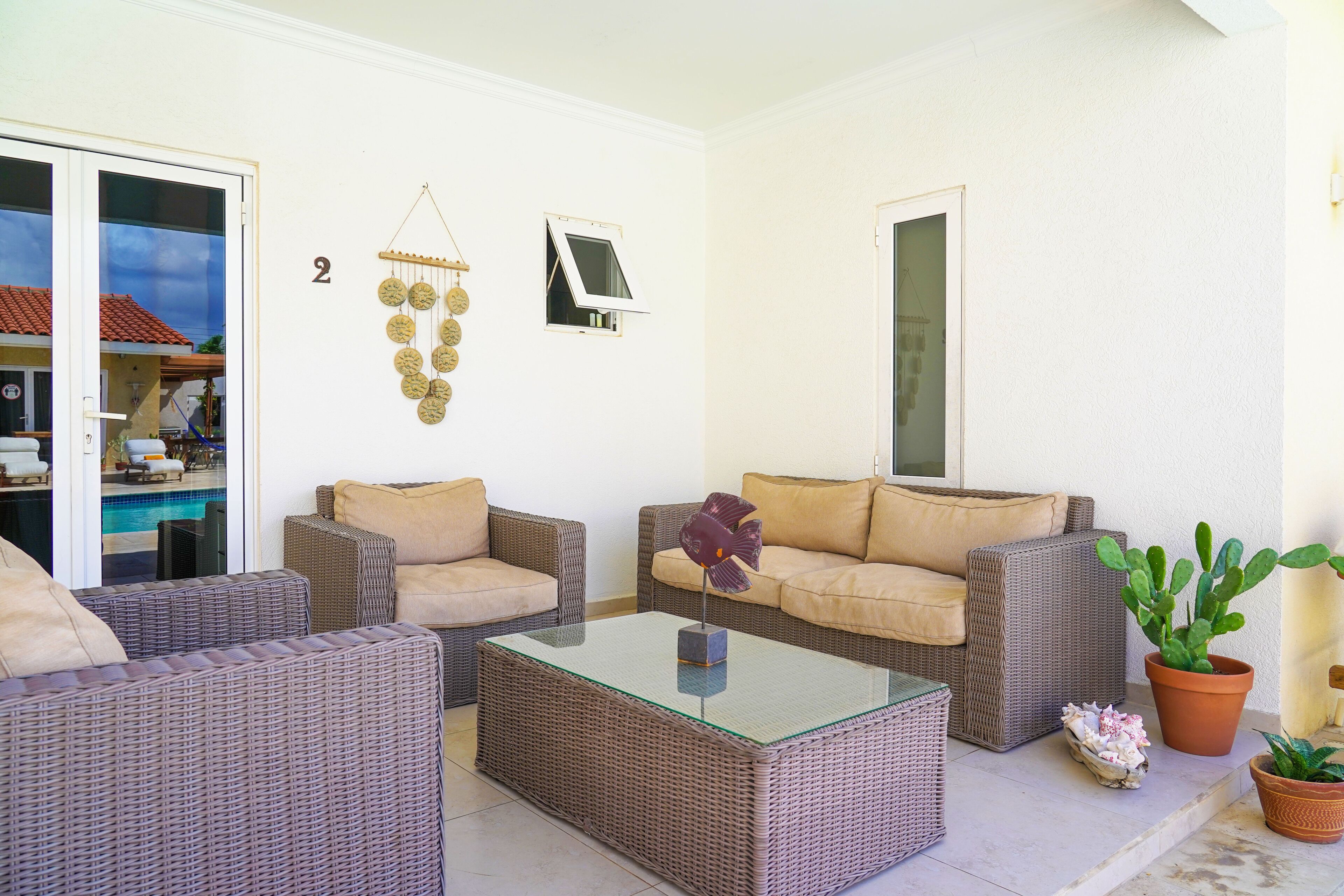 Um pátio ensolarado com móveis de vime, uma mesa com tampo de vidro e um vaso de planta.