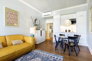 Uma sala de estar moderna com um sofá amarelo, uma mesa de jantar com cadeiras e uma área de cozinha com armários brancos e um lustre.