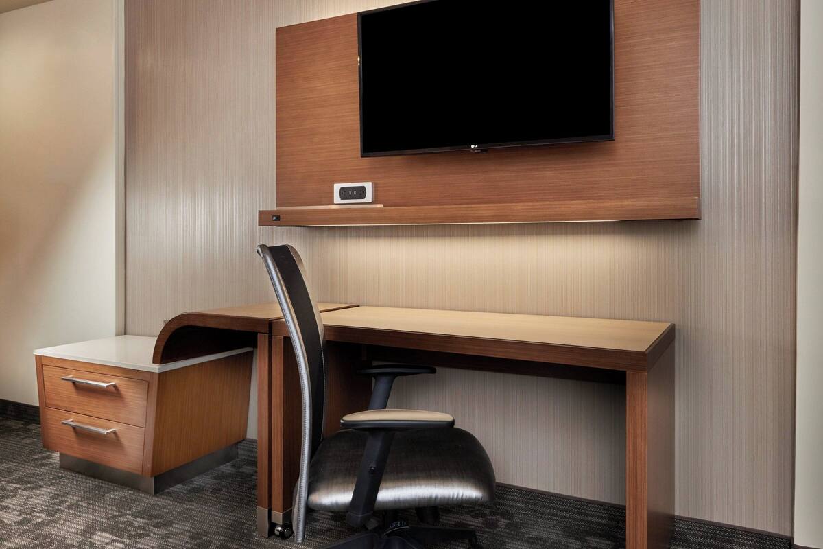 Quarto de hotel com escrivaninha de madeira, uma cadeira, televisão fixada na parede e uma pequena cômoda.