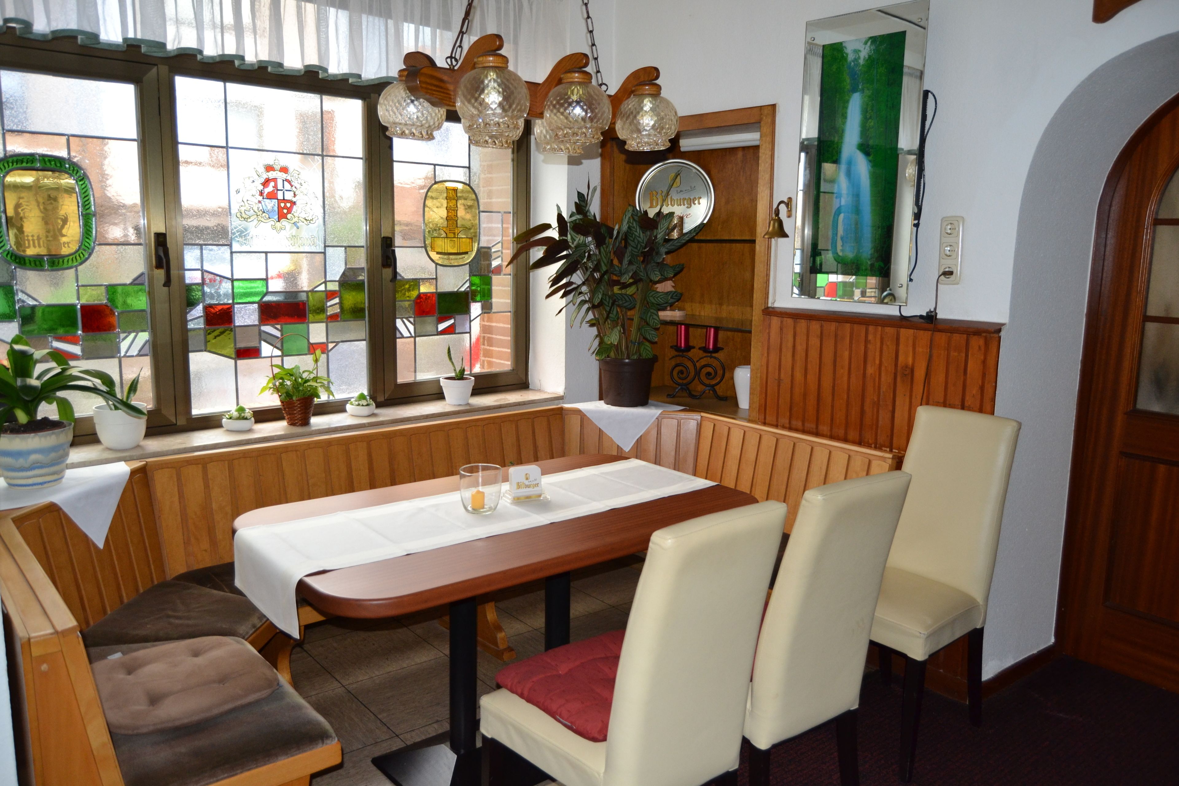 Uma área de refeições acolhedora com um banco de madeira, uma mesa e cadeiras. Há plantas em vasos e uma janela de vitrais.