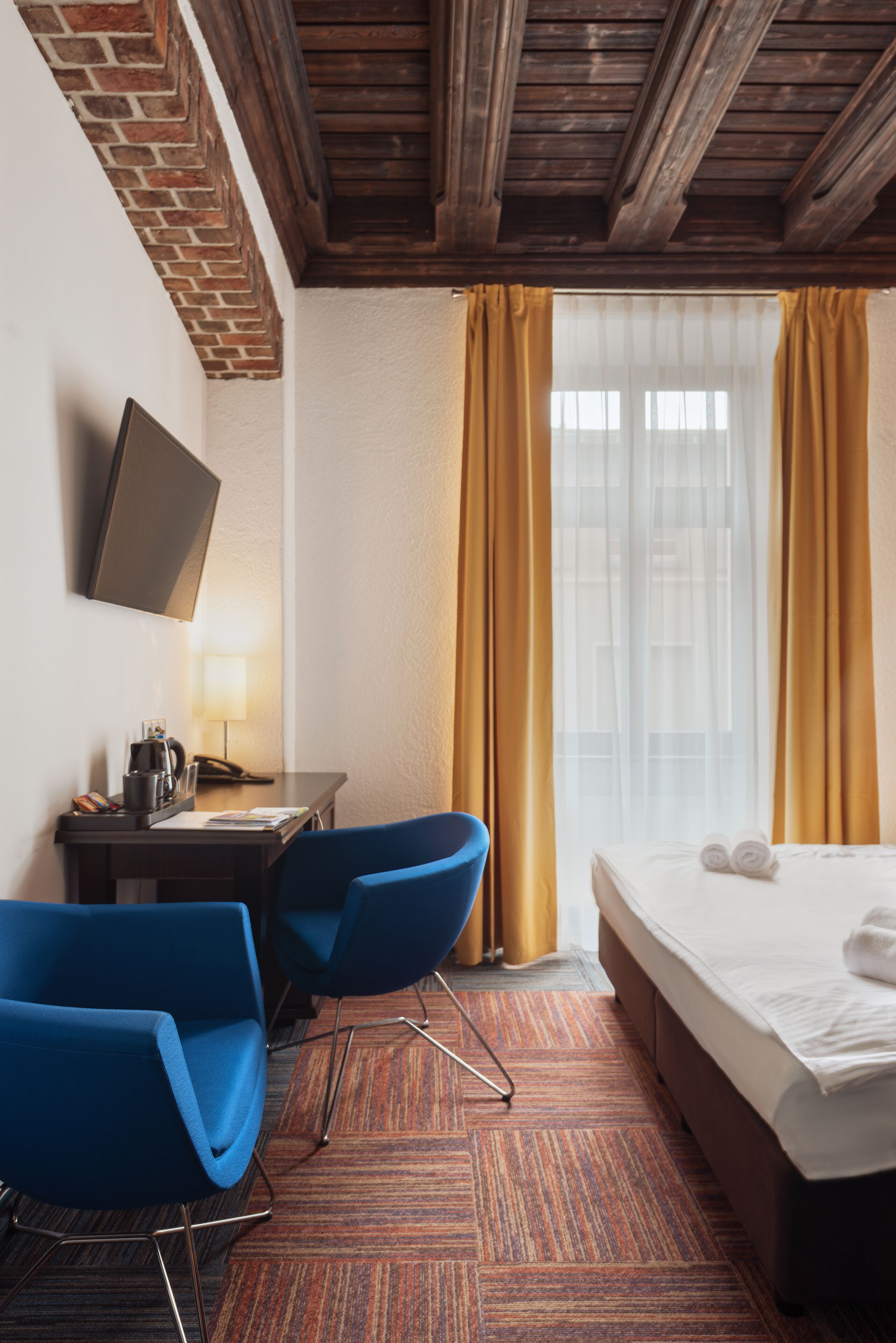 Quarto de hotel com cama, duas cadeiras azuis, escrivaninha com abajur, televisão na parede e janela com cortinas amarelas.