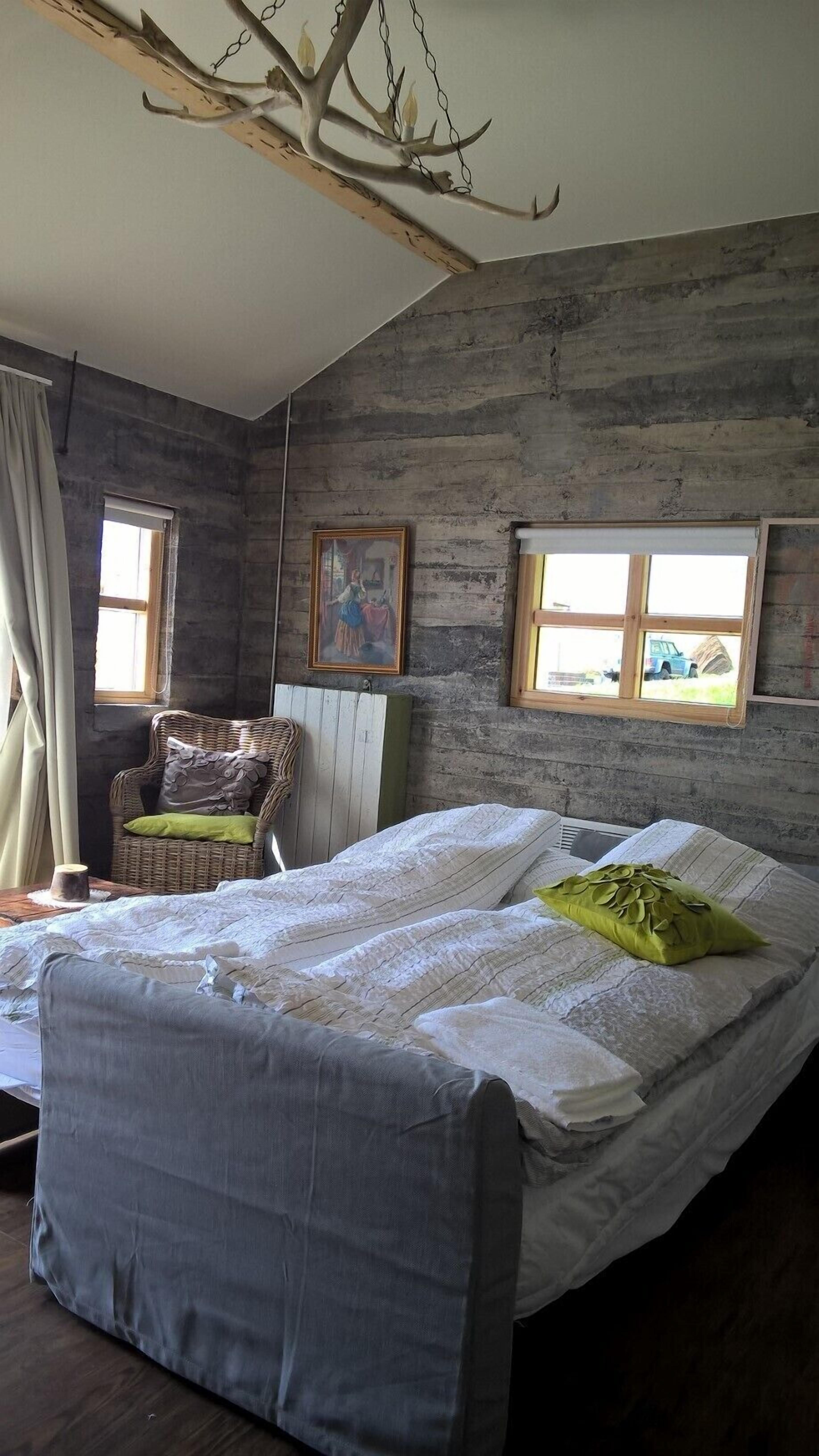 Quarto com cama, uma cadeira de vime, um quadro e uma janela com vista.