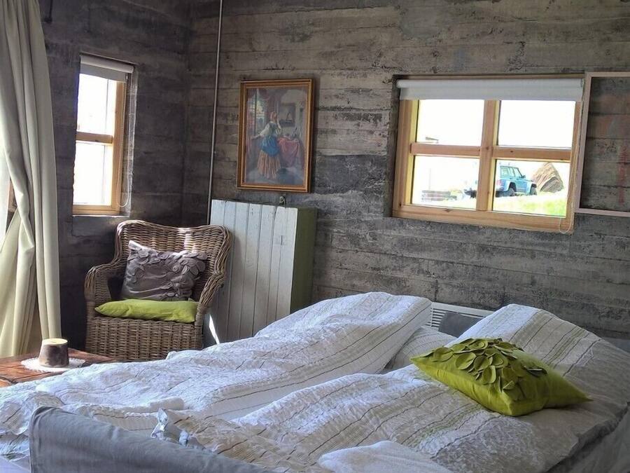 Quarto com cama, uma cadeira de vime, um quadro e uma janela com vista.