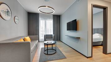 Quarto de hotel moderno com sofá cinza, mesa de centro redonda, televisão na parede e cama com cabeceira cinza.