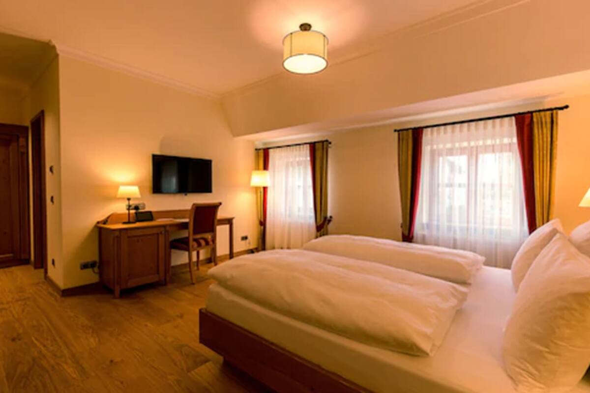 Quarto de hotel com uma cama grande, uma mesa com cadeira, televisão e cortinas nas janelas.
