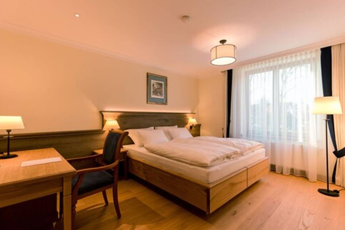 Quarto de hotel com cama de madeira, uma escrivaninha com cadeira, mesa de cabeceira, luminária e janela com cortinas.