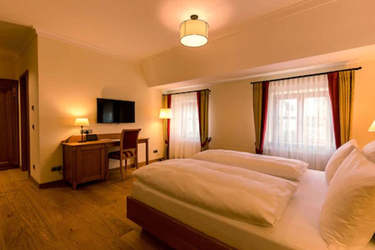 Quarto de hotel com cama, escrivaninha, cadeira, televisão e cortinas.