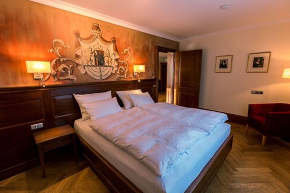 Quarto de hotel com uma cama grande, cabeceira de madeira, duas luminárias de cabeceira, uma pequena mesa de apoio e uma cadeira vermelha.