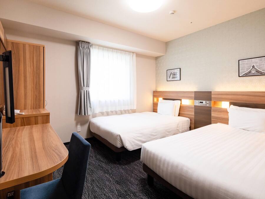 Quarto de hotel com duas camas, uma escrivaninha, uma cadeira, janela com cortinas, cabeceira com iluminação embutida e um quadro na parede.