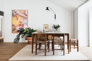 Uma área de refeições com mesa e cadeiras de madeira, uma planta em vaso e um quadro na parede.