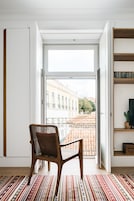 Quarto com vista para um pátio, uma cadeira de madeira, um tapete listrado e uma estante com livros e um cacto.