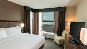 Quarto de hotel com uma cama grande, uma mesa com TV, uma cadeira e vista para a água pela janela.