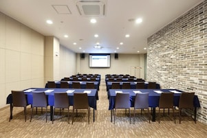 Uma sala de conferências com fileiras de mesas e cadeiras, uma tela de projetor e uma parede de tijolos.