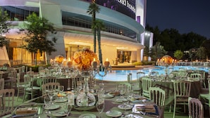 Uma área de refeições ao ar livre com mesas preparadas para um banquete, piscina ao fundo e um edifício de hotel com uma placa indicando "elite world hotels".