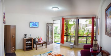 Sala de estar com TV, móveis de madeira, uma mesa pequena com um laptop e vista para um jardim através de grandes janelas.