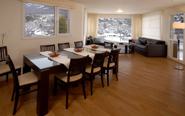 Uma sala de jantar com mesa de vidro e cadeiras de madeira, um sofá, e vista para uma paisagem nevada através de grandes janelas.
