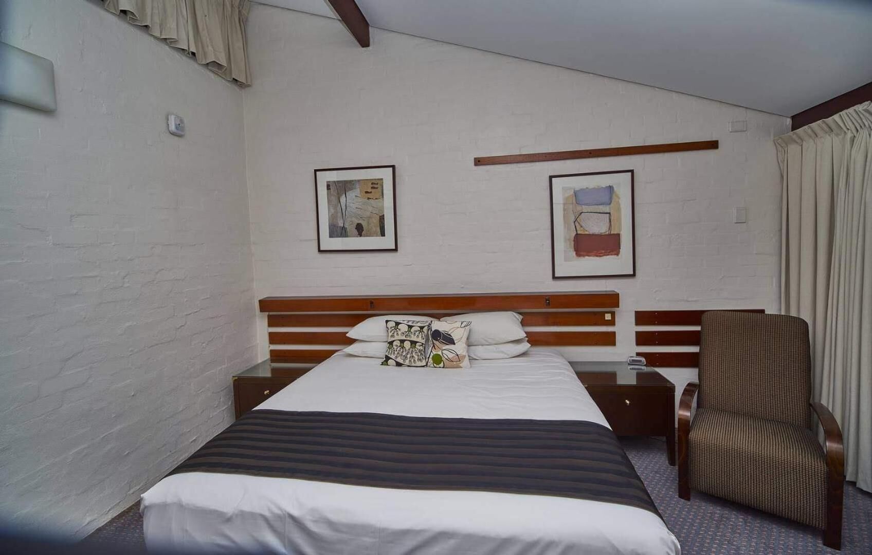 Quarto de hotel com uma cama grande, cabeceira de madeira, duas obras de arte emolduradas na parede, uma cadeira e uma mesa de cabeceira com um telefone.