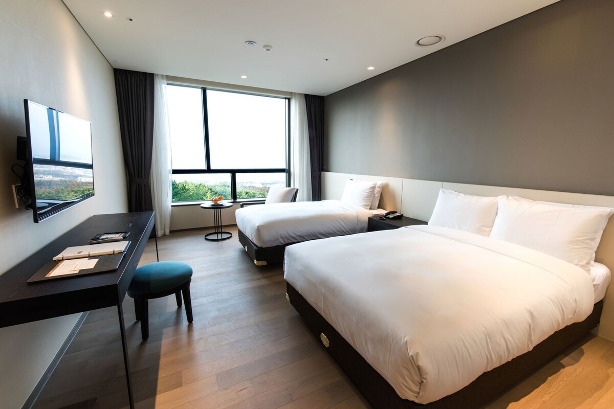 Quarto de hotel com duas camas, uma escrivaninha, uma cadeira, televisão e janela com vista.