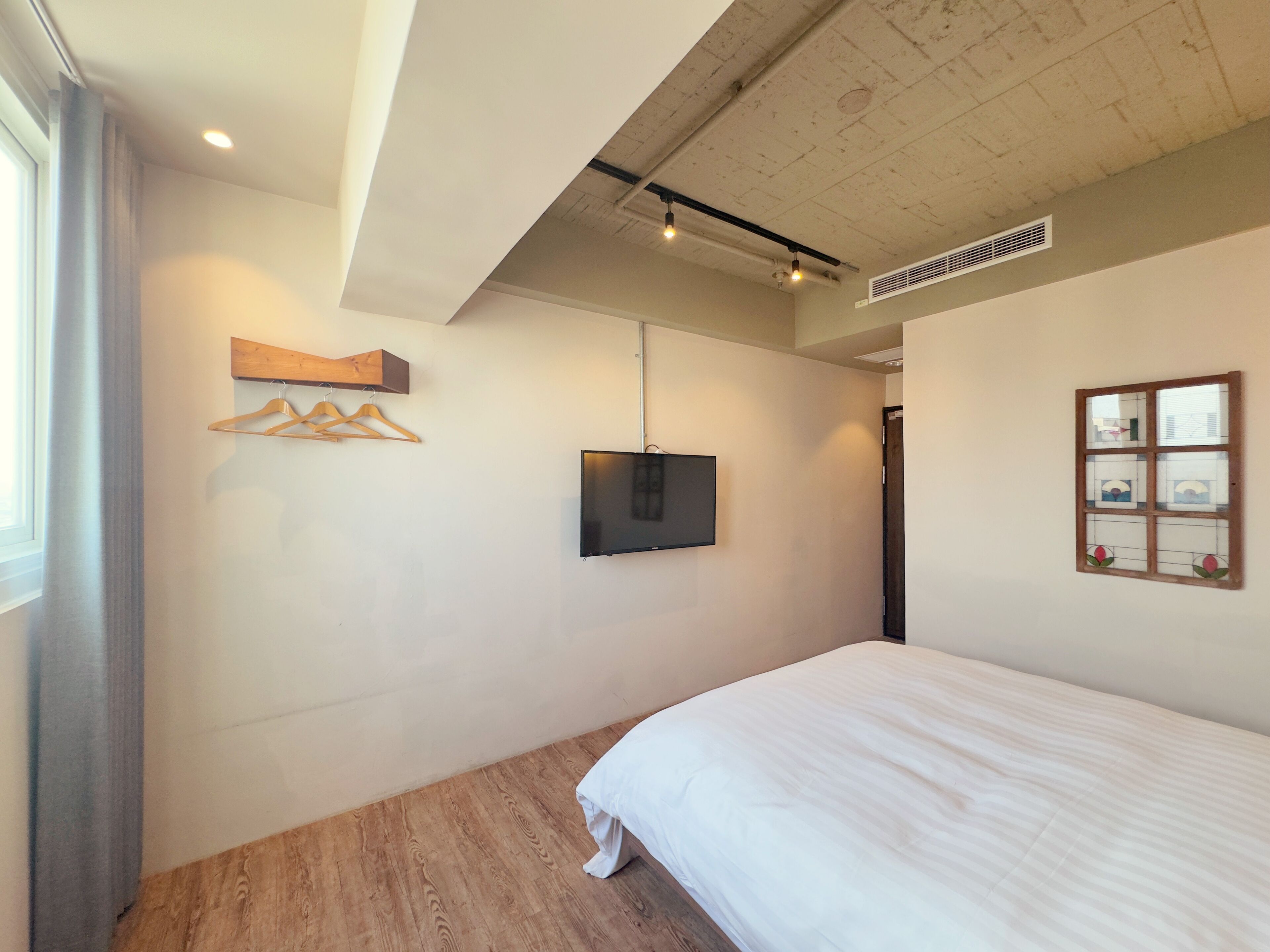 Quarto com cama, televisão fixada na parede, cabides de madeira e janela.