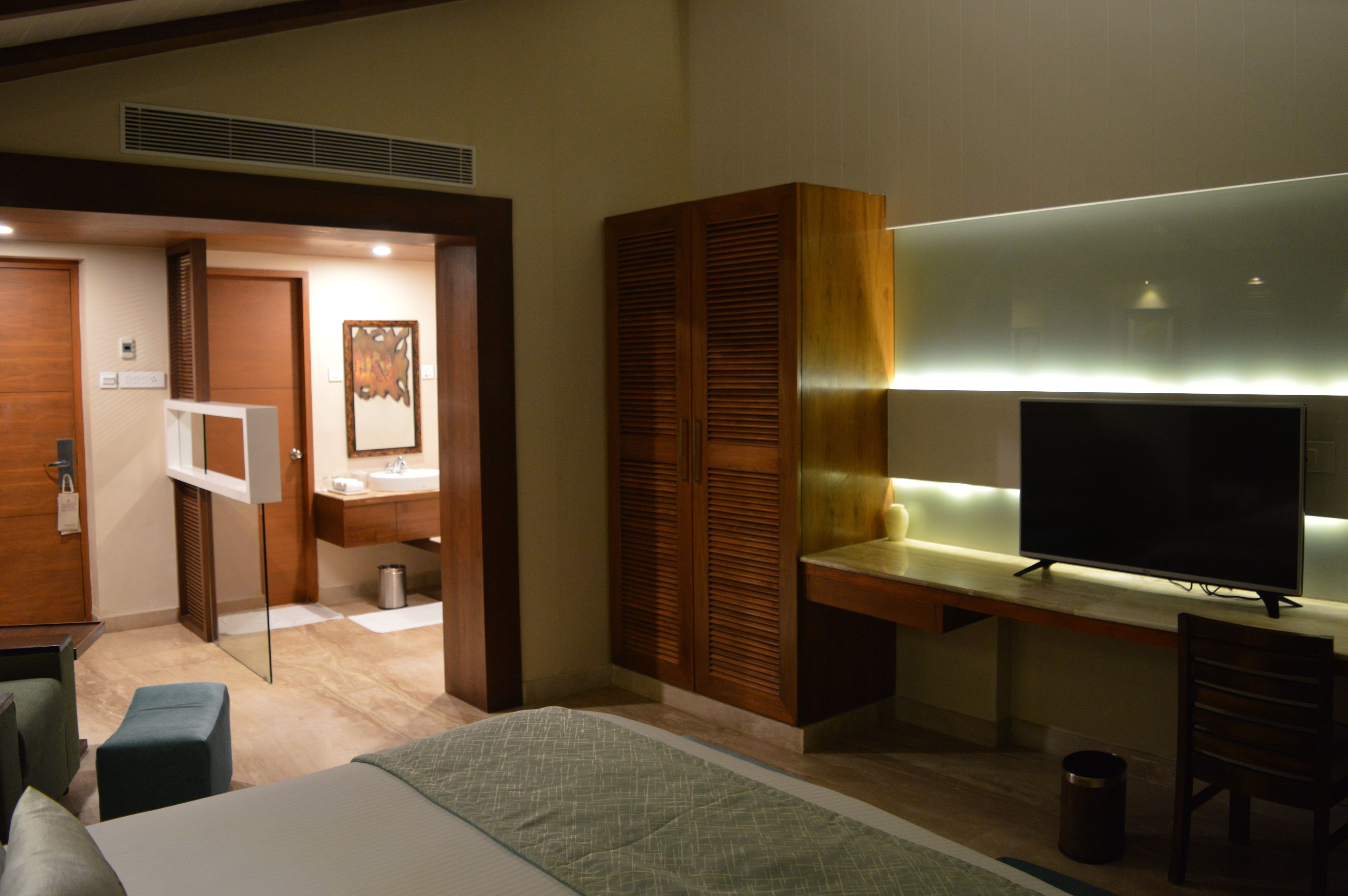 Quarto de hotel com cama, escrivaninha com televisão, espelho e guarda-roupa de madeira.