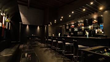 Um espaço de bar com iluminação suave, vários taburetes e mesas, um balcão com diversos frascos e copos, e um design moderno no teto.