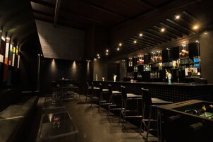 Um espaço de bar com iluminação suave, vários taburetes e mesas, um balcão com diversos frascos e copos, e um design moderno no teto.
