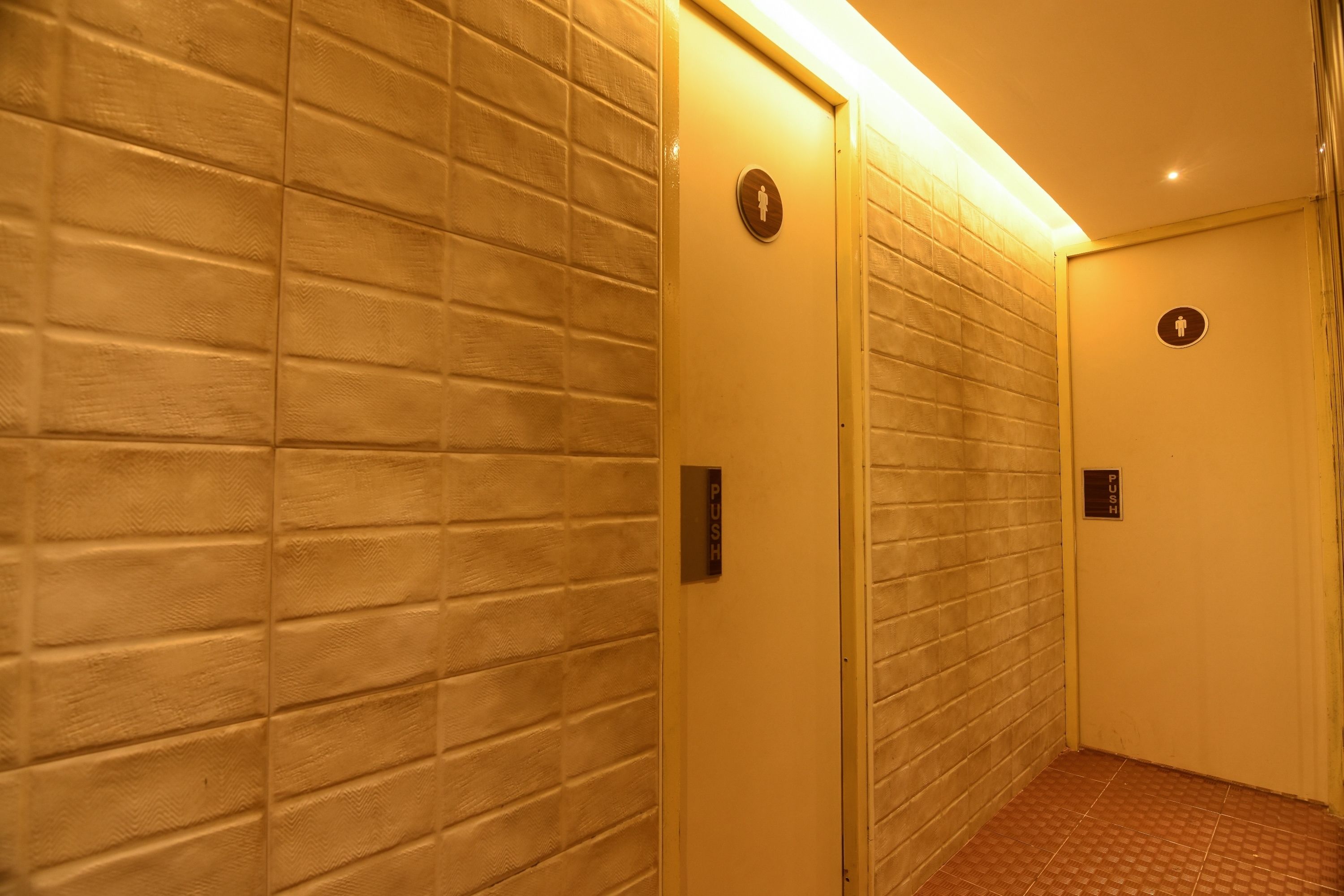 Corredor com parede texturizada e chão em azulejos. Há duas portas com botão de pressão e uma placa indicando um banheiro.