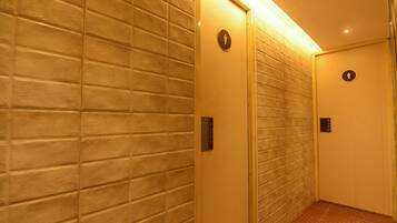 Corredor com parede texturizada e chão em azulejos. Há duas portas com botão de pressão e uma placa indicando um banheiro.