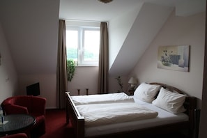 Quarto com duas camas, uma cadeira vermelha, uma mesinha redonda e uma janela com cortinas.