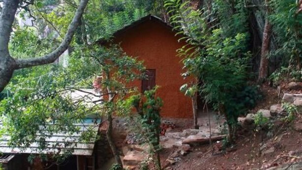 Um edifício rústico, térreo, com telhado inclinado, rodeado por vegetação densa e árvores.