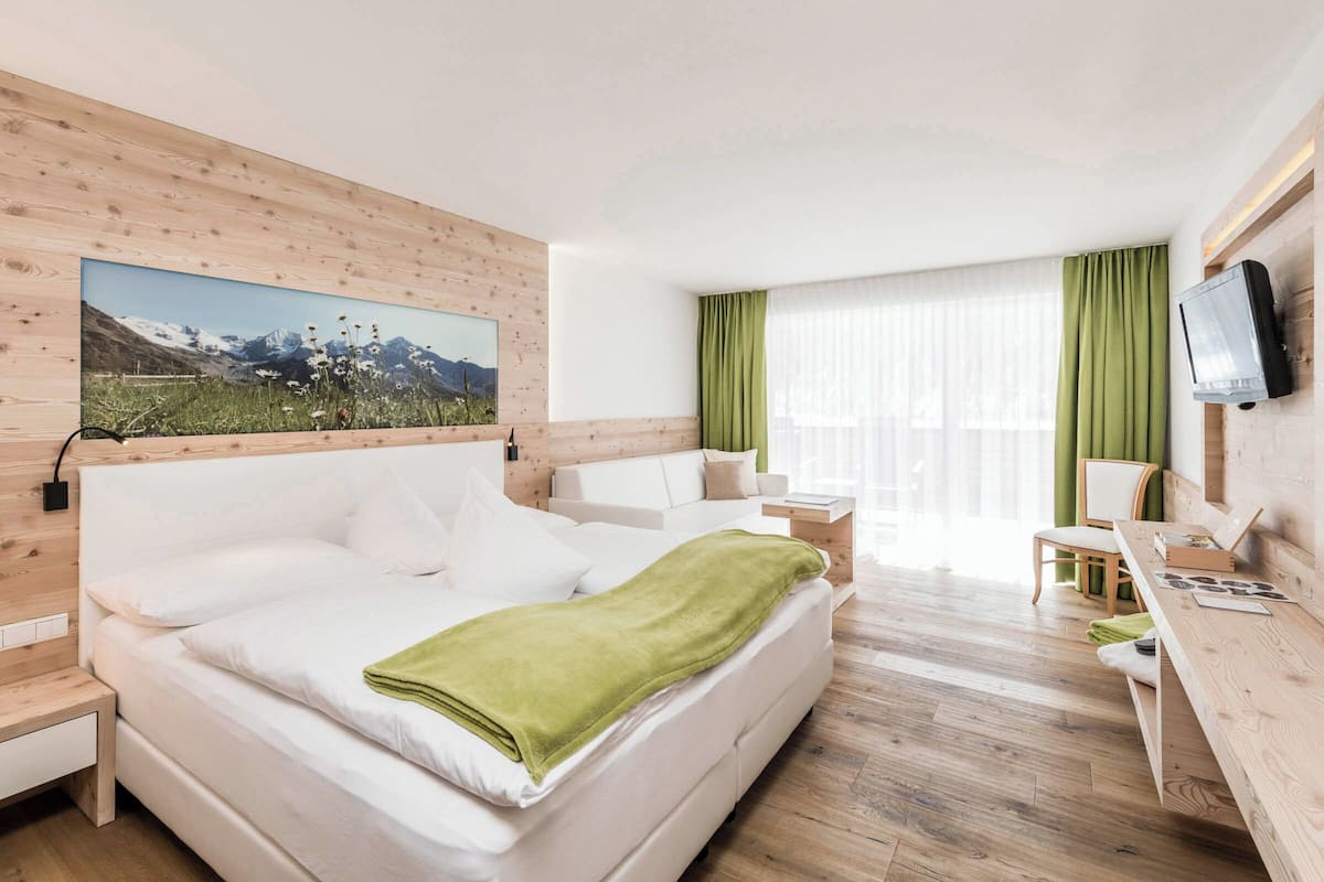 Quarto de hotel com cama, escrivaninha, cadeira, televisão e um quadro na parede.